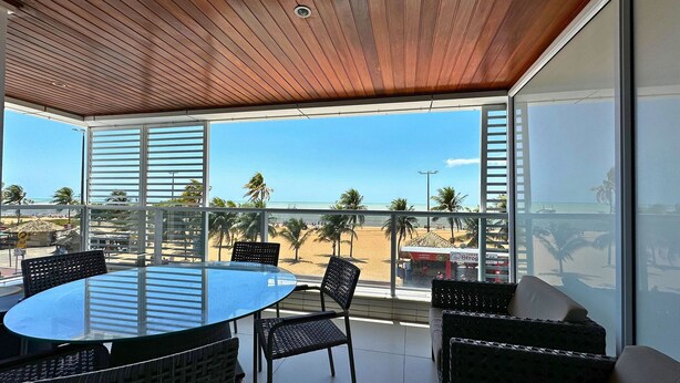 Uma varanda com teto de madeira, mesa de vidro e cadeiras, com vista para uma praia com palmeiras e céu limpo.