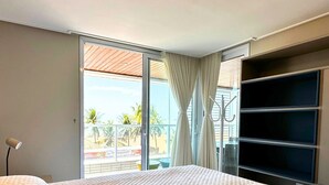 Quarto de hotel com cama, escrivaninha, cadeira, janela com cortinas e varanda com vista para palmeiras.