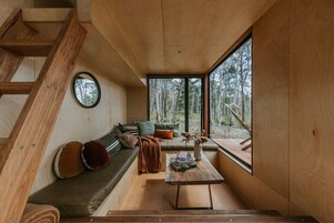 Interior acolhedor de uma cabana com escada de madeira, um sofá com almofadas, mesa de centro e janelas amplas com vista para uma floresta.