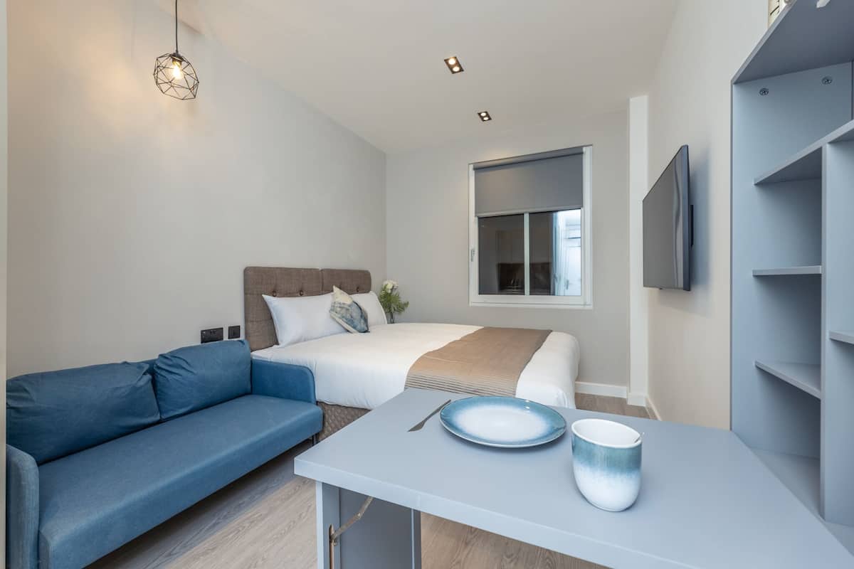 Quarto de hotel moderno com cama, sofá azul, pequena mesa com prato e caneca, TV na parede e janela com vista.