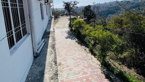 Uma trilha leva a um edifício com janelas protegidas por grades, um aparelho de ar condicionado e vista para uma paisagem montanhosa.