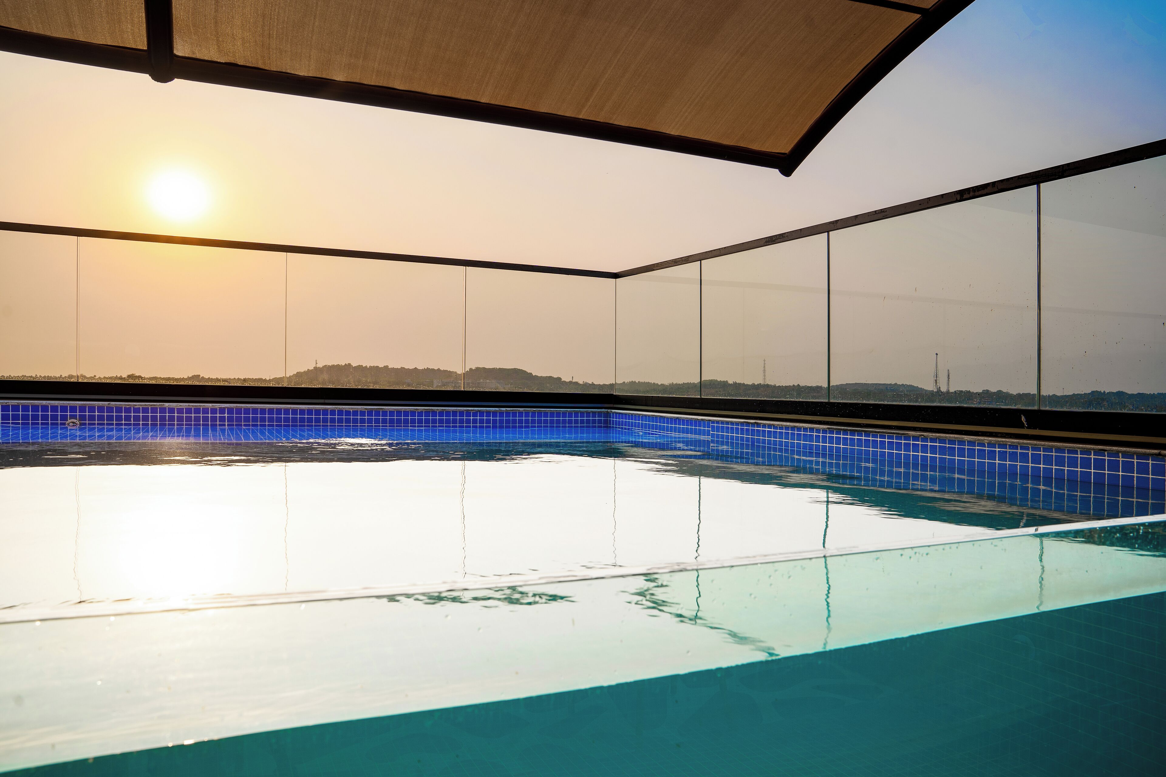 Uma piscina interna com paredes de vidro transparente, vista para o pôr do sol e piso revestido de azulejos.