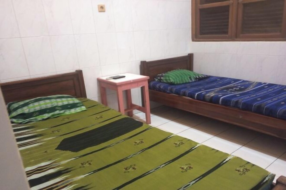Quarto pequeno e simples com duas camas de solteiro, uma mesa pequena e uma janela.
