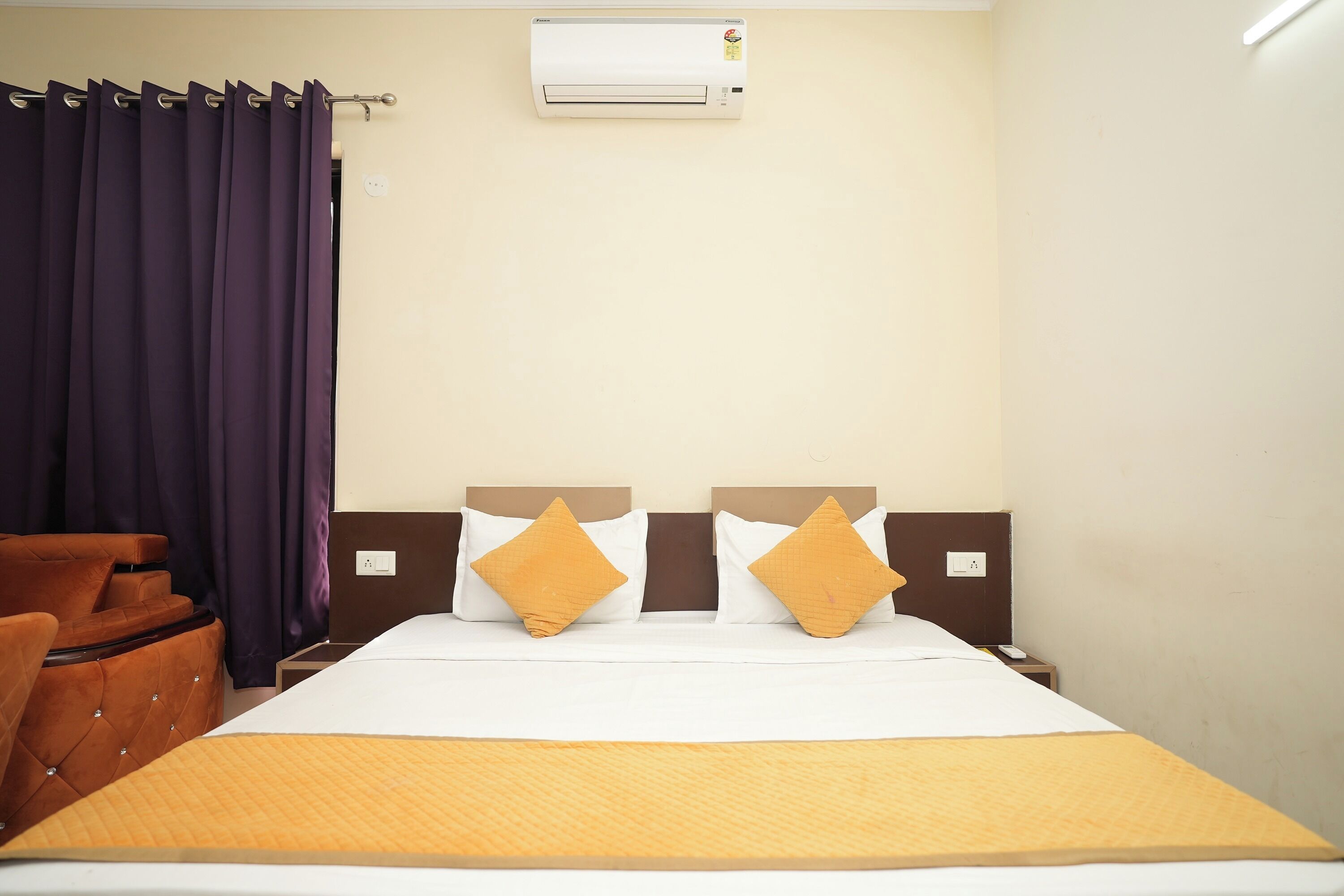 Quarto de hotel com cama, duas almofadas, mesa de cabeceira, cadeira e ar condicionado.