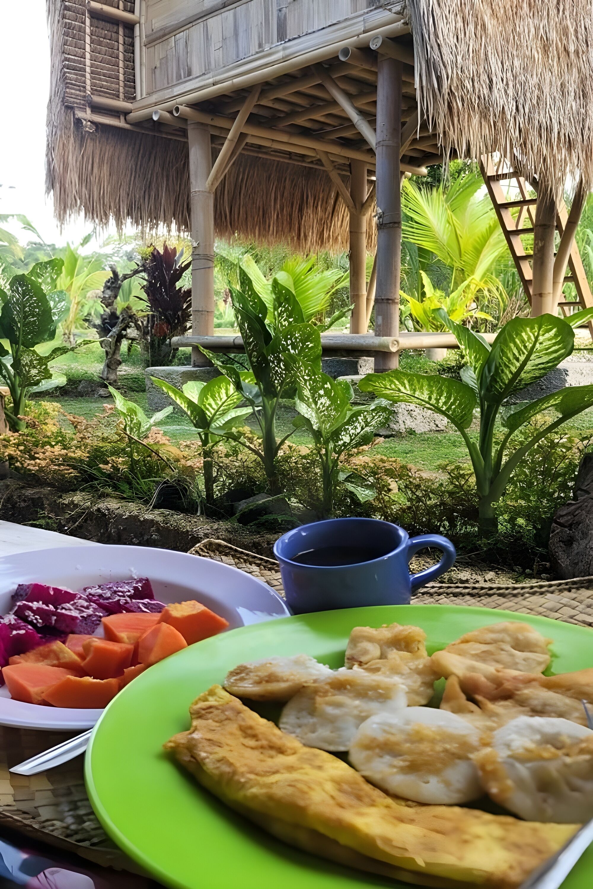 Uma mesa de jantar com um prato de comida, uma xícara azul e uma construção com telhado de palha ao fundo.