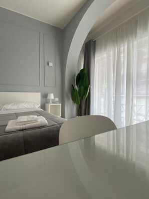 Quarto de hotel moderno com cama, mesa de cabeceira, luminária e uma janela grande com cortinas.
