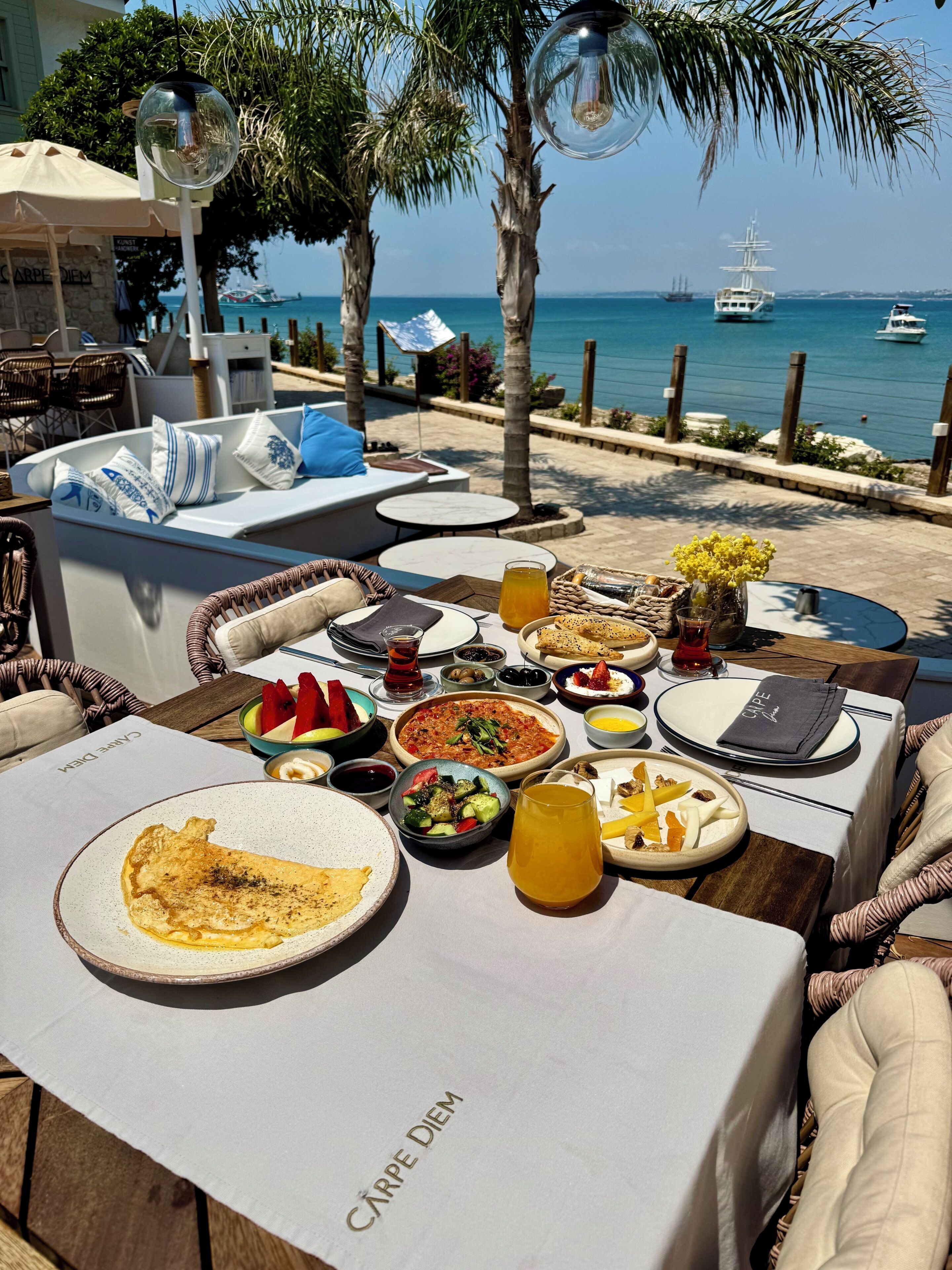 Mesa posta com itens de café da manhã, incluindo ovos, pão e frutas, com vista para a costa, barcos e palmeiras.