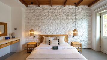 Quarto com parede de pedra, vigas de madeira no teto, cama com roupa de cama branca, duas mesas de cabeceira com abajures e uma mesinha de cabeceira com um abajur.