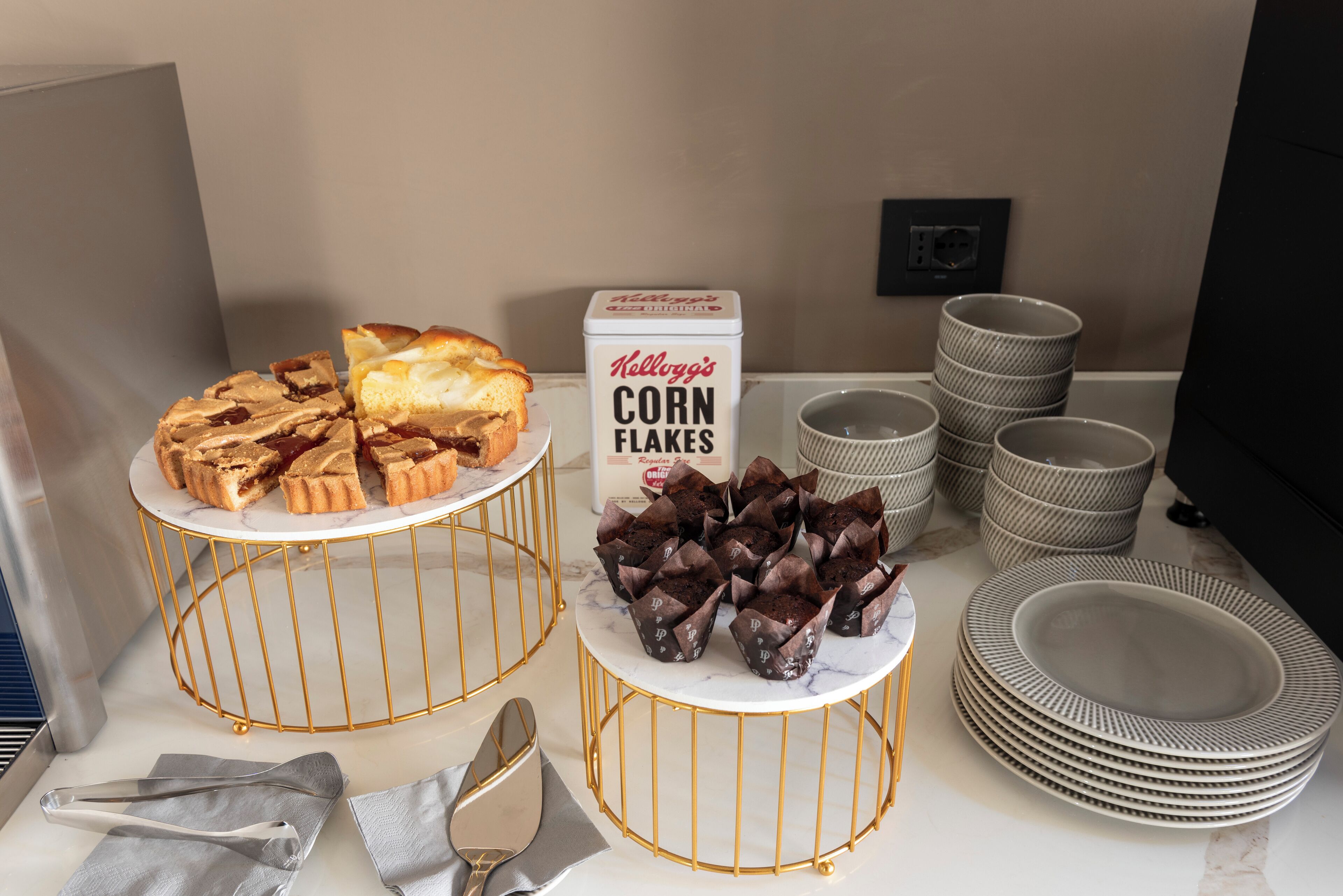 Uma bancada de cozinha com uma caixa de Kellogg's Corn Flakes, pilhas de pratos e um bolo em suporte de arame dourado.