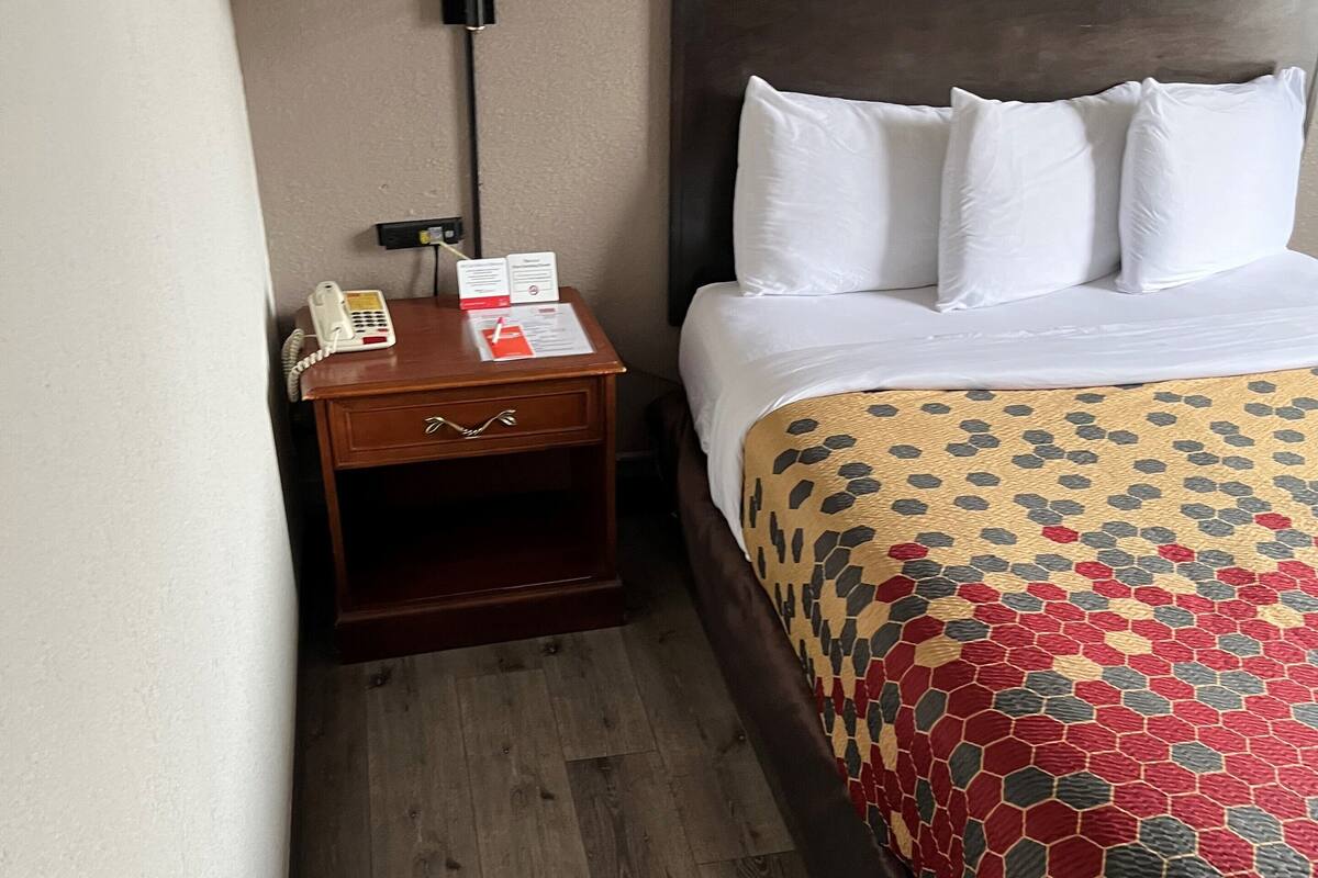 Quarto de hotel com chão de madeira, cama com roupa de cama branca e manta estampada, mesa de cabeceira com abajur, telefone e pequena gaveta.