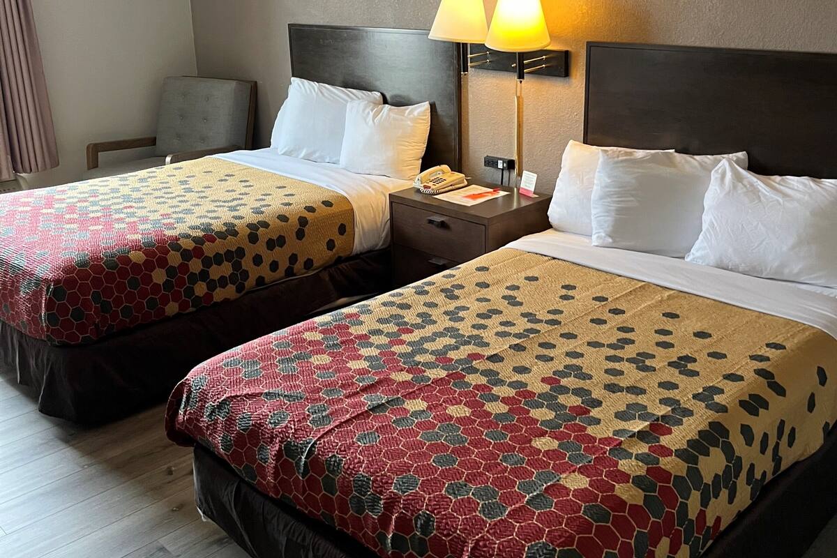 Quarto de hotel com duas camas, cada uma com colcha estampada, mesa de cabeceira com abajur e uma cadeira.