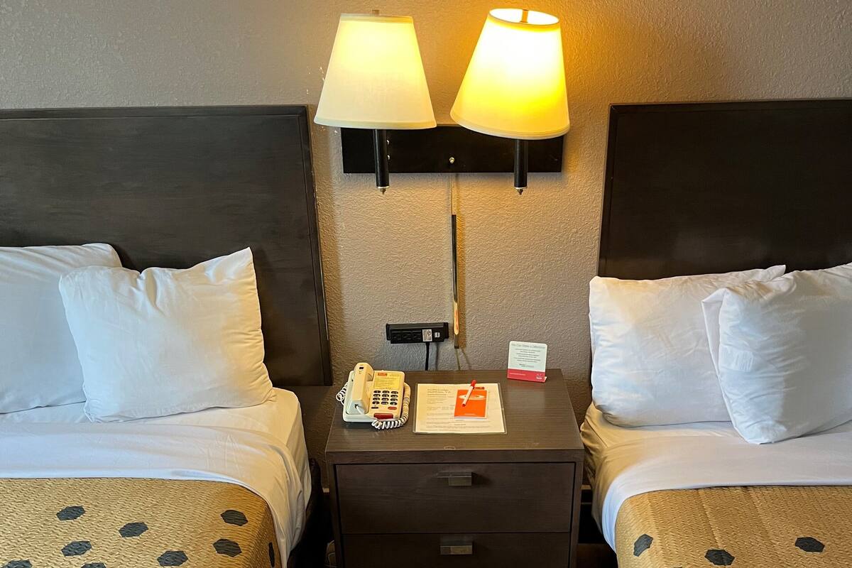 Quarto de hotel com duas camas, cada uma com roupa de cama branca e uma colcha com padrão. Há uma mesinha de cabeceira entre as camas com um telefone, um bloco de notas e uma placa vermelha. O quarto possui piso de madeira e parede texturizada.
