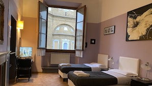 Quarto de hotel com duas camas, uma televisão, uma janela com vista para um edifício e um quadro na parede.