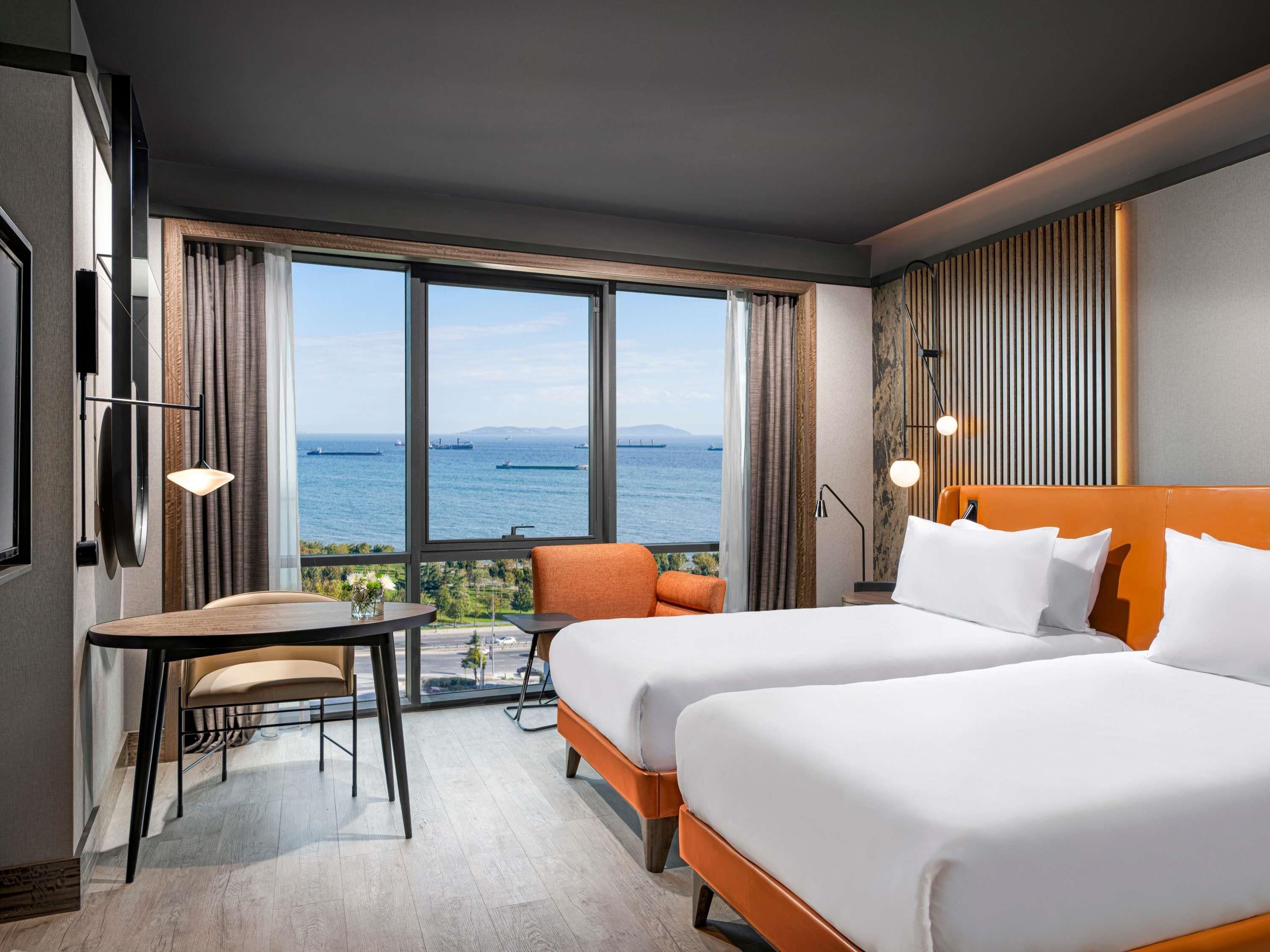 Quarto de hotel com duas camas, uma mesa pequena, uma cadeira e vista para o mar.
