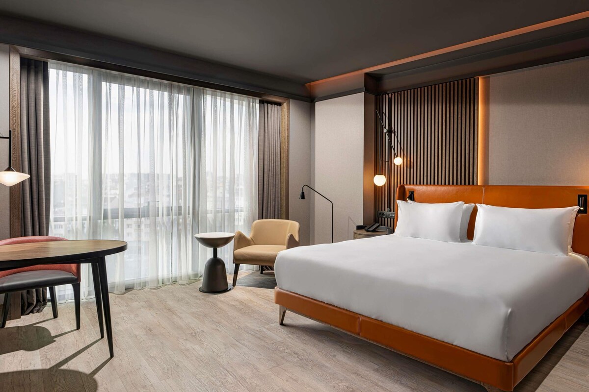 Quarto de hotel com uma cama grande, uma escrivaninha, uma cadeira e vista para a cidade através de cortinas leves.