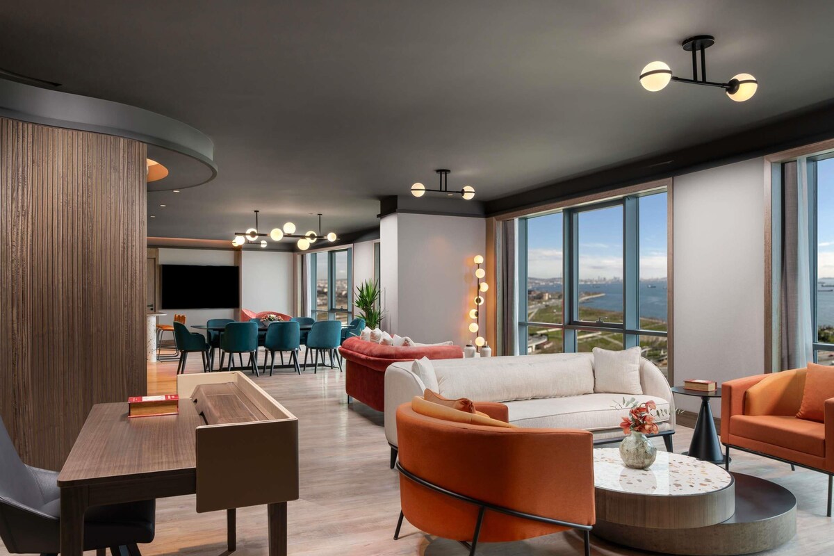 Uma sala de estar moderna com uma parede de destaque em madeira, uma área de jantar com cadeiras cor turquesa e uma grande janela com vista para a cidade.