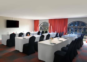 Uma sala de conferências com várias mesas, cadeiras e um grande janelão com cortinas vermelhas.