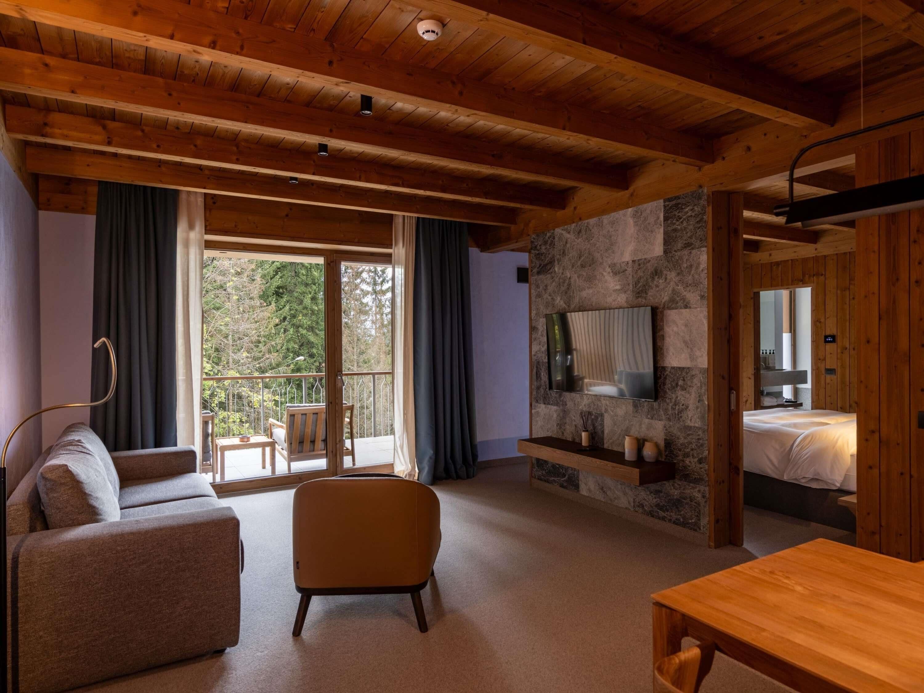 Um quarto acolhedor com teto de madeira, uma janela grande com cortinas, um sofá, uma cadeira, uma TV fixada em uma parede de pedra e uma cama com cabeceira de madeira.