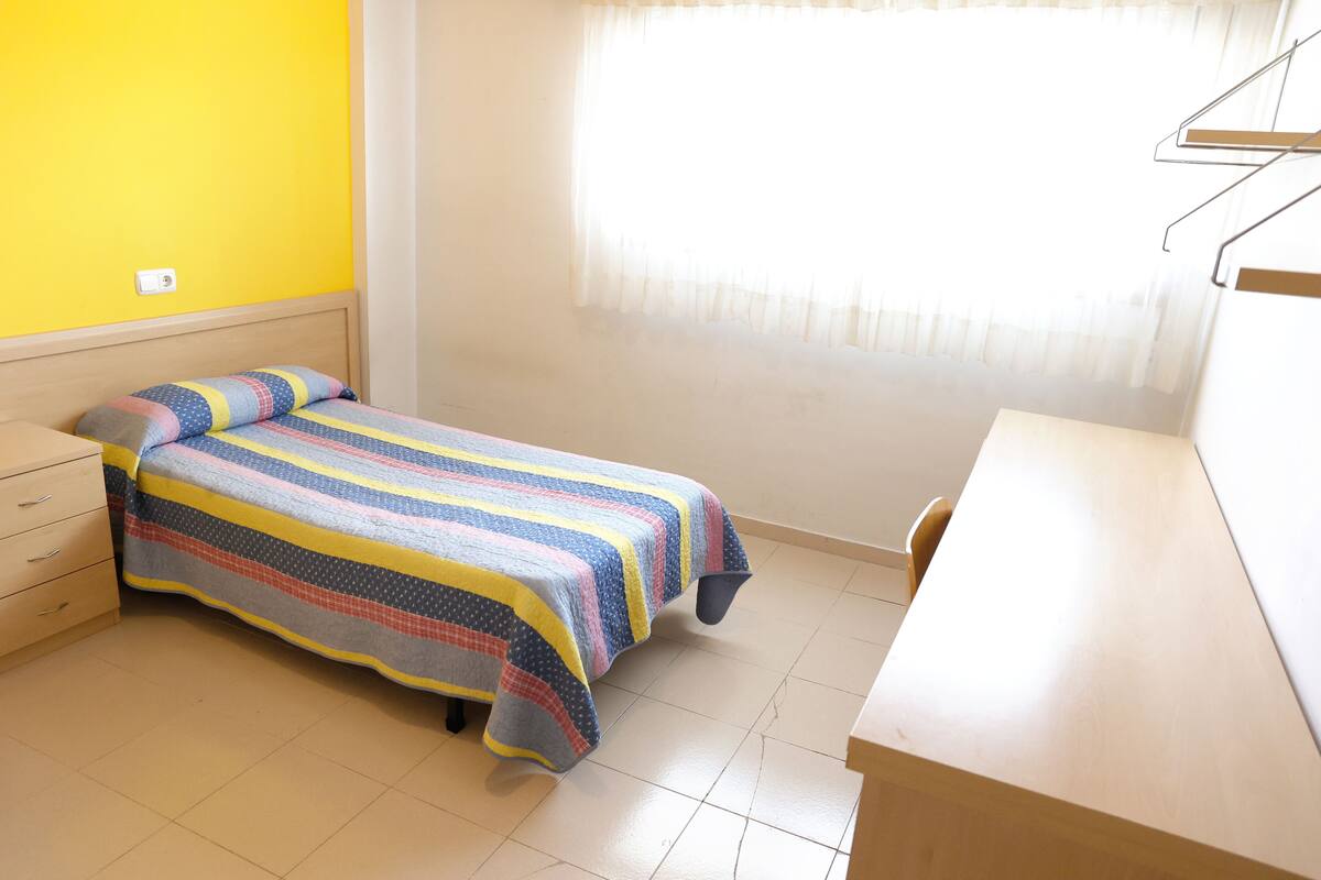 Um quarto com cama, escrivaninha e janela com cortinas.