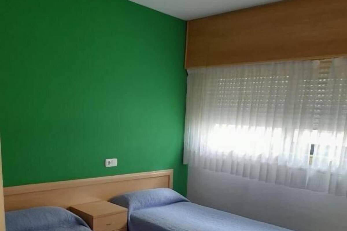 Quarto com duas camas de solteiro, cabeceira de madeira, janela com cortinas finas e parede verde.