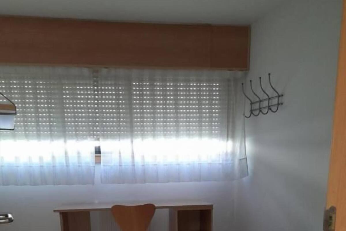 Quarto pequeno e bem iluminado com uma cama, uma escrivaninha e uma cadeira.