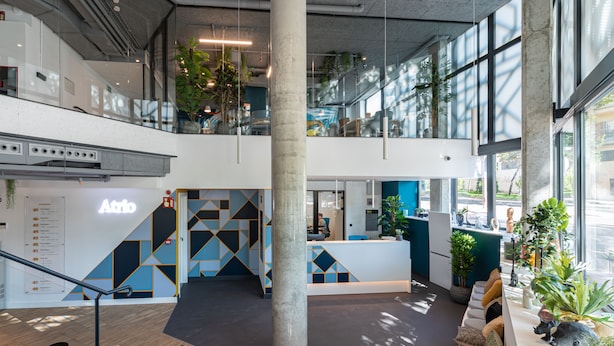 Uma área de recepção moderna com design geométrico na parede, um pilar central e um espaço de estar com plantas.