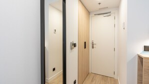 Quarto de hotel moderno com piso de madeira, porta branca com puxador preto e um pequeno armário de madeira à direita.