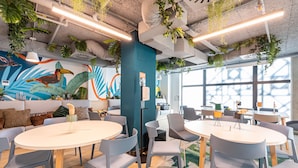 Um espaço de escritório moderno com mesas redondas, cadeiras cinzas e um mural de aves tropicais.