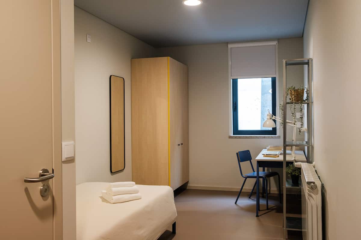 Quarto de hotel pequeno com uma cama, um guarda-roupa, uma secretária e uma cadeira, além de uma janela.