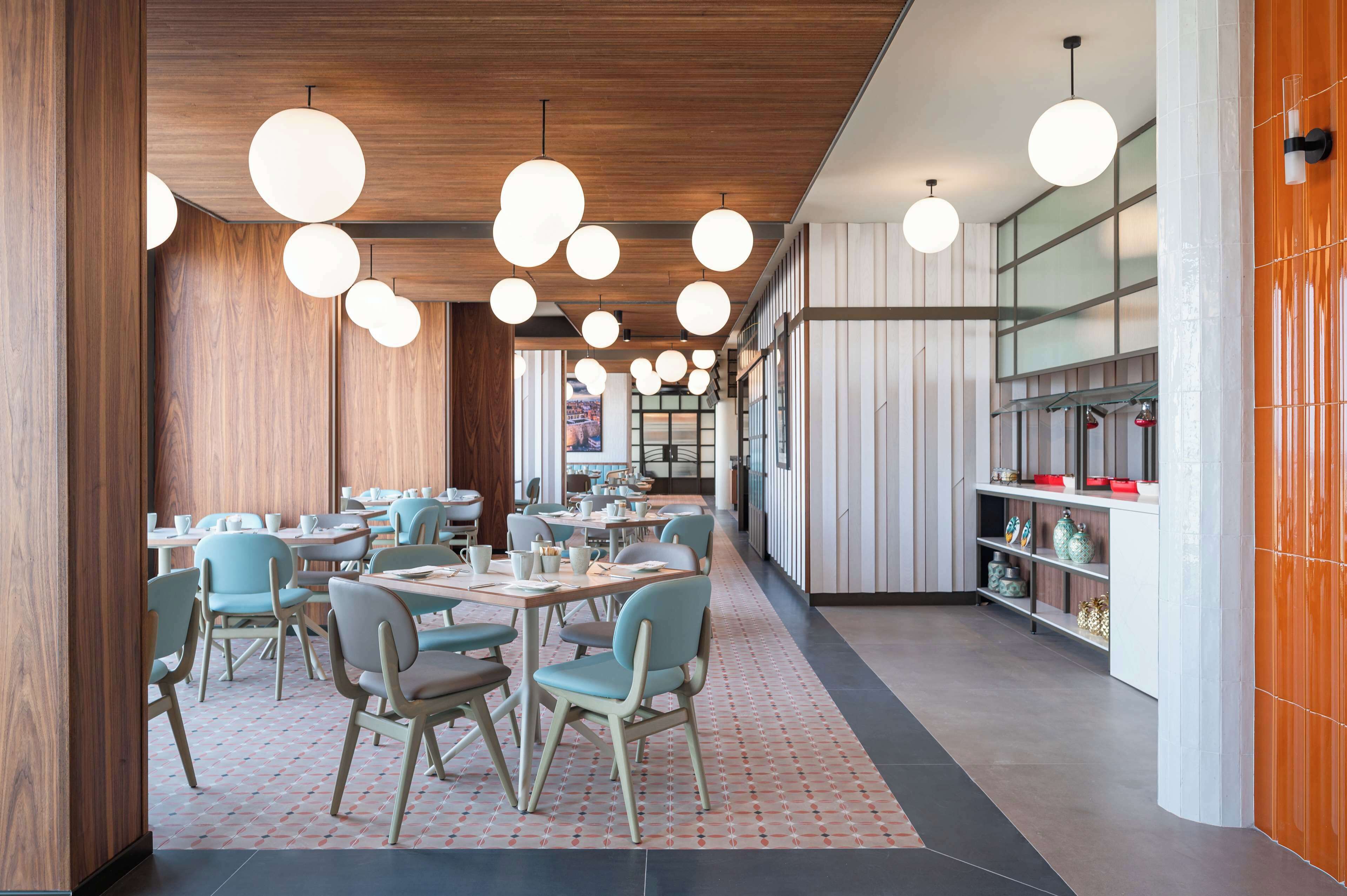 Um restaurante moderno com painéis de teto em madeira, luminárias pendentes redondas e piso estampado. Mesas e cadeiras dispostas para refeições.