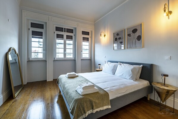 Quarto com cama, mesa de cabeceira, espelho, duas janelas e obras de arte na parede.