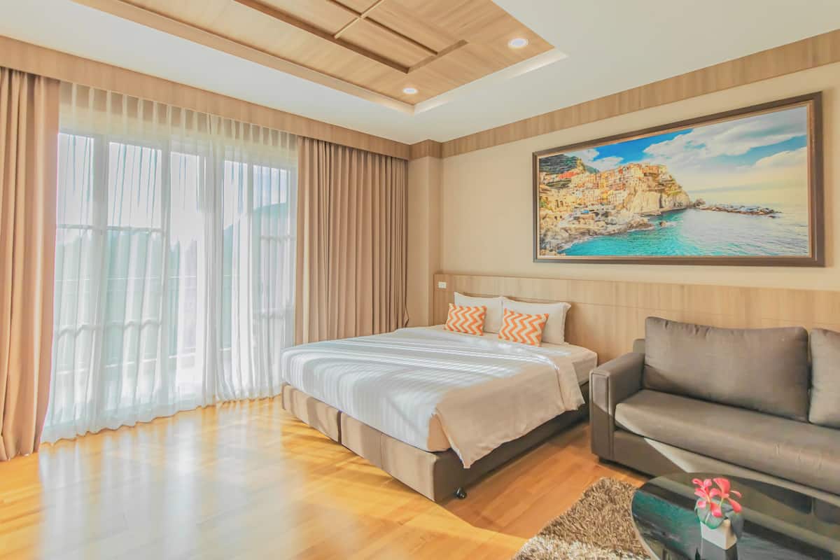 Quarto de hotel com uma cama grande, um sofá, uma pintura de paisagem costeira e piso de madeira.