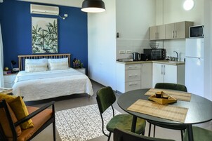 Quarto de hotel compacto com uma cama, uma pequena cozinha compacta, área de refeições com mesa e cadeiras, e uma parede azul de destaque.