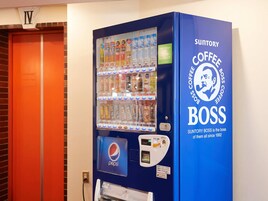 Máquina de vendas com exterior azul e logotipo da Pepsi, localizada em um corredor com uma porta vermelha.