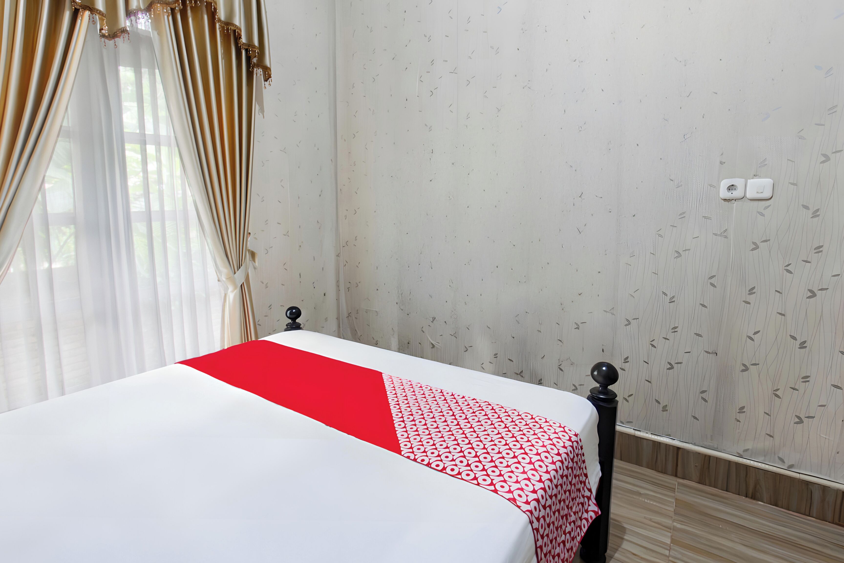 Uma cama com colcha branca e vermelha, piso de madeira, parede com padrão de folhas e janela com cortinas.