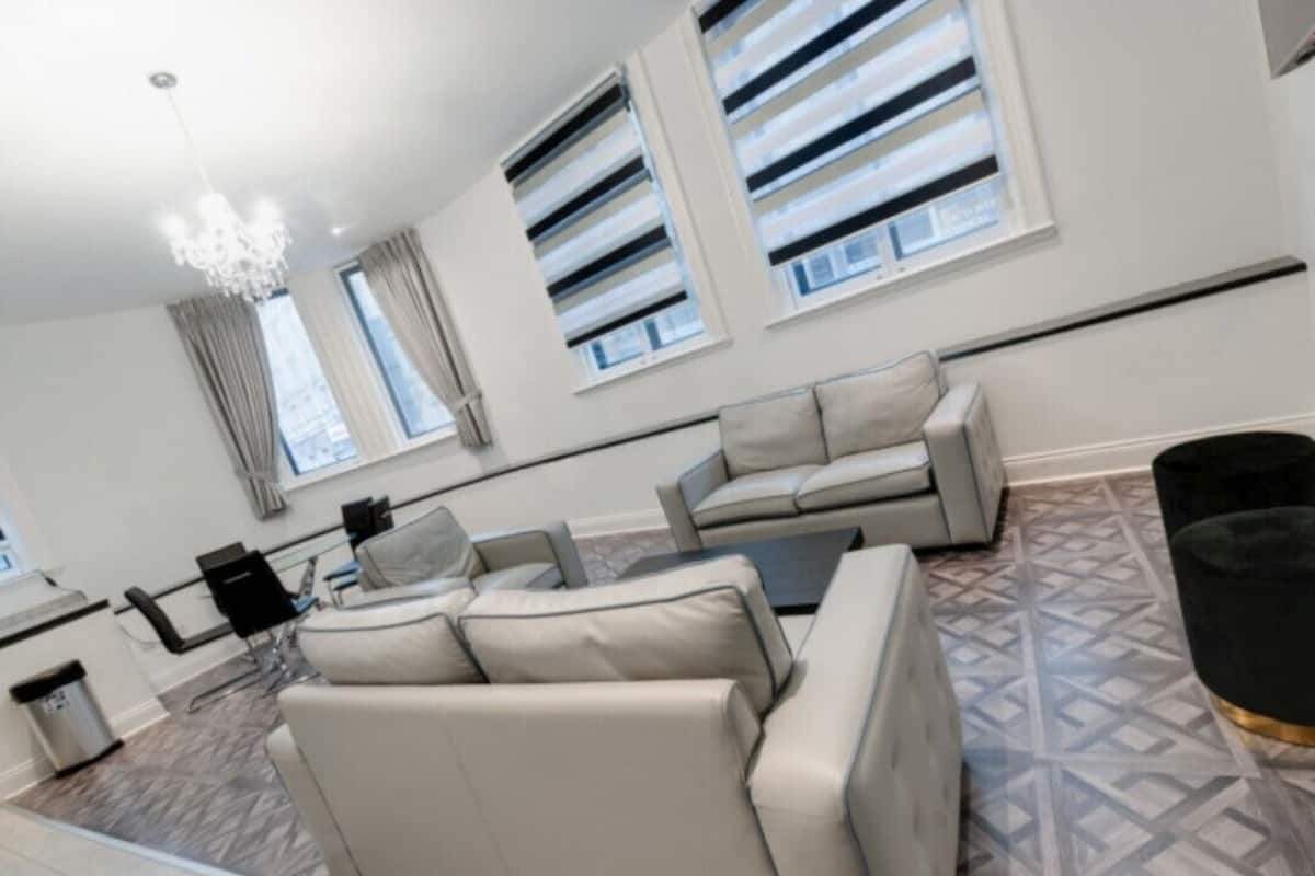 Uma sala de estar moderna com um sofá, uma mesa de centro e um lustre. Há janelas com persianas, uma lixeira e um piso estampado.