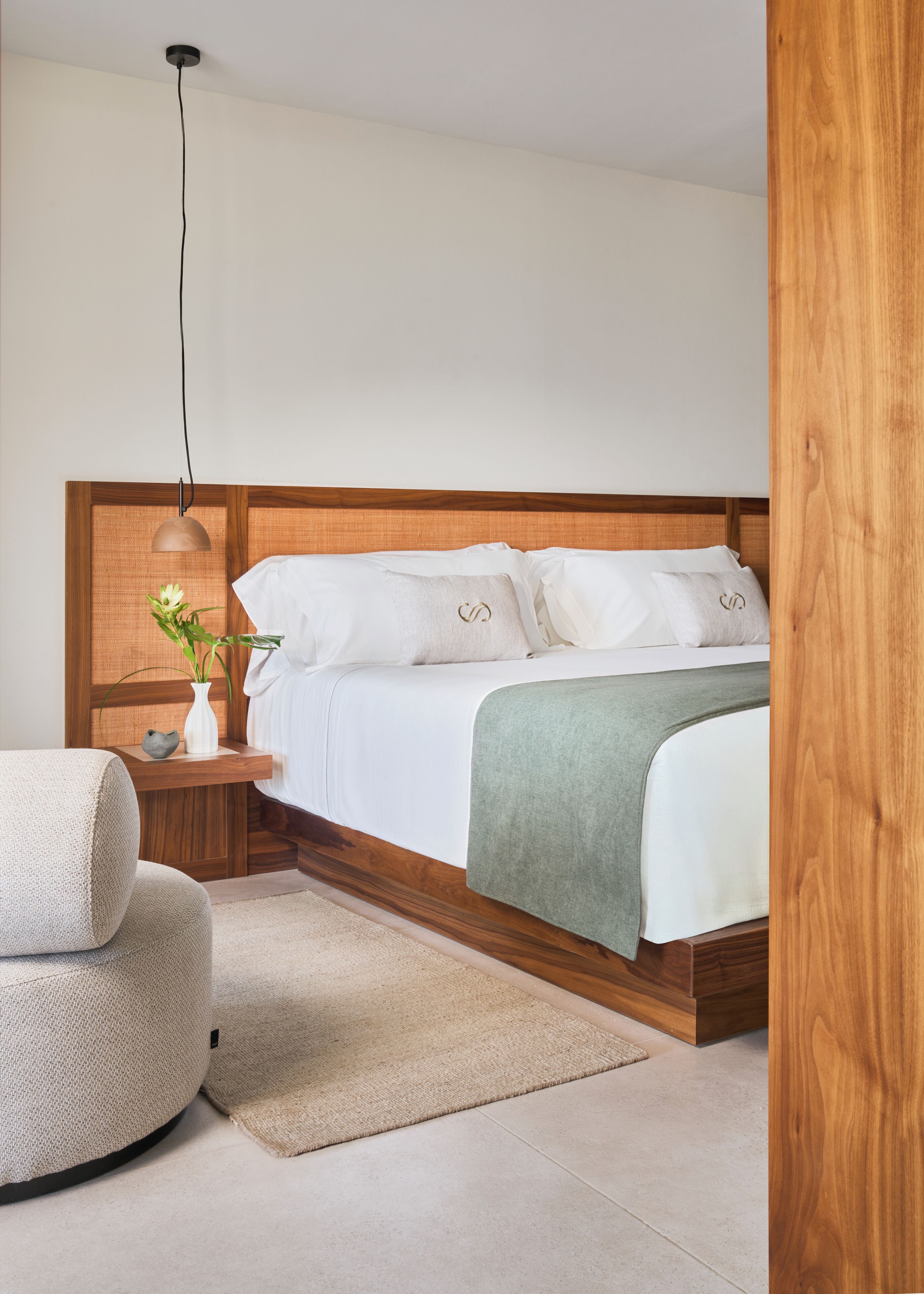 Quarto com cabeceira de madeira, cama com lençóis brancos, manta verde, mesa de cabeceira com um vaso e uma planta, e um abajur suspenso.