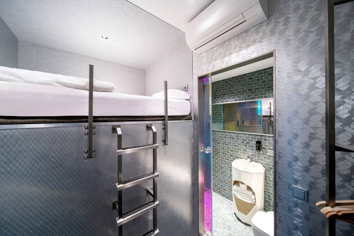 Quarto de hotel compacto com beliche, escada de metal, banheiro com parede revestida em azulejo e uma pequena janela.
