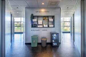 Estação de autoatendimento com toalhas, lixeira e contentor de reciclagem num corredor com grandes janelas.