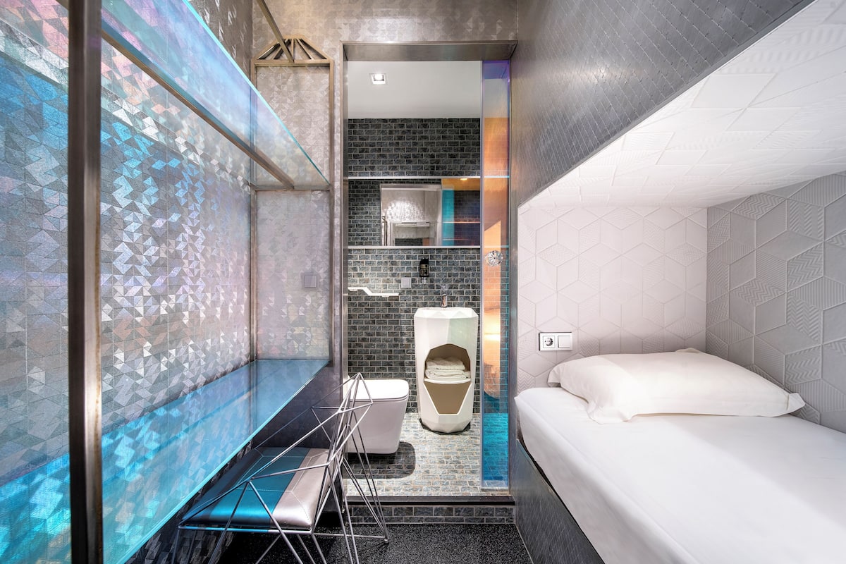 Quarto de hotel compacto com cama, banheiro pequeno e um design de parede geométrico singular.
