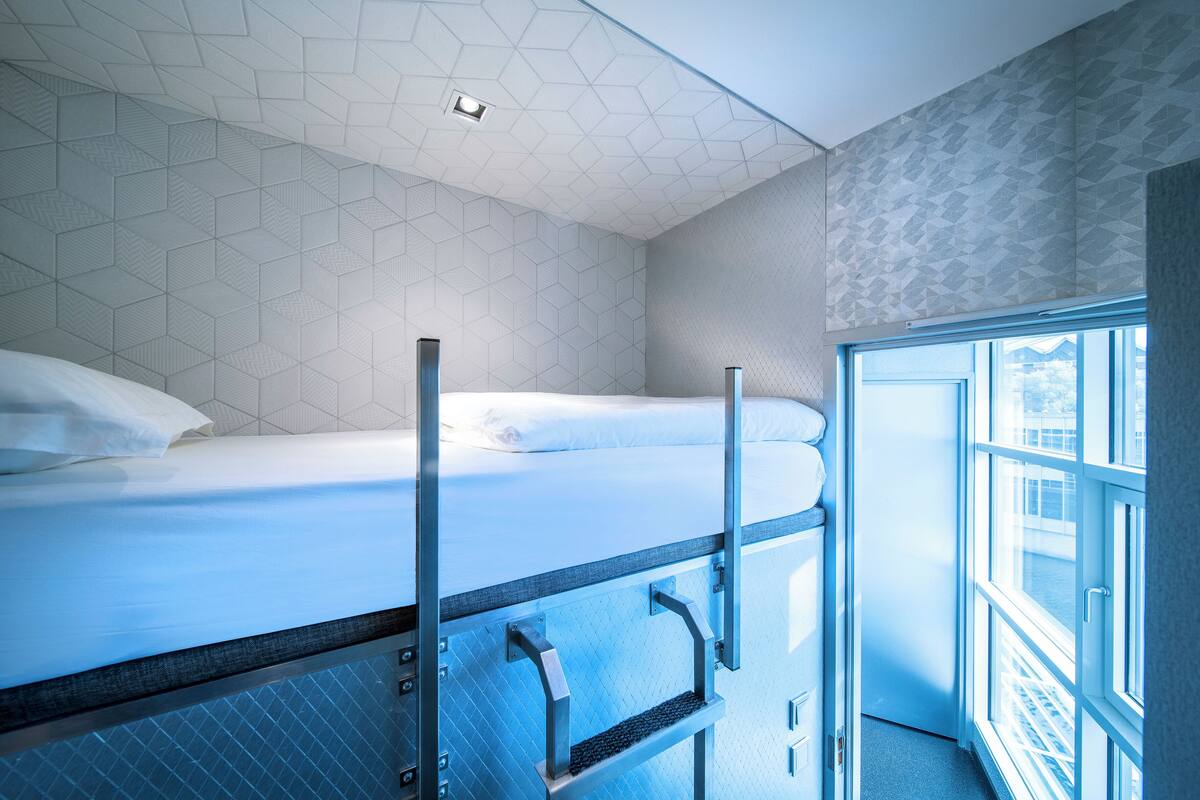 Quarto de hotel moderno com beliche, paredes com padrão geométrico, uma janela e uma escada de metal.