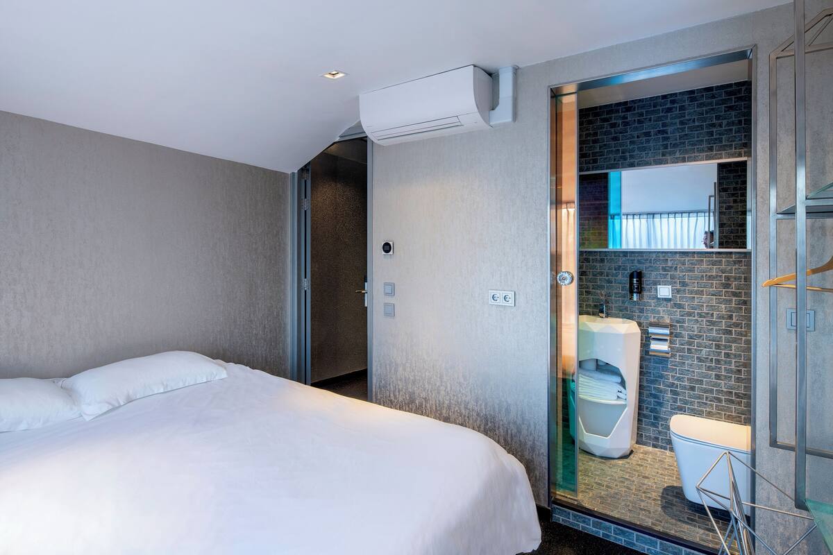 Quarto de hotel com cama, banheiro envidraçado e parede de tijolos.
