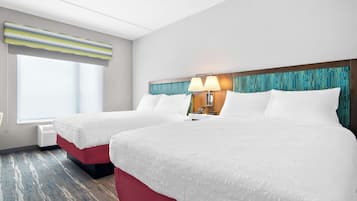 Quarto de hotel com duas camas, uma janela grande com persiana listrada, uma luminária fixada na parede e um tapete estampado.