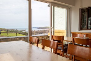 Uma área de refeições com mesa e cadeiras, uma janela grande com vista para o mar e falésias, e uma parede de pedra.
