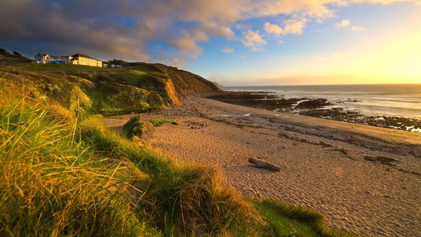 Paisagem costeira com área gramada, praia de areia e um edifício no topo de um penhasco.