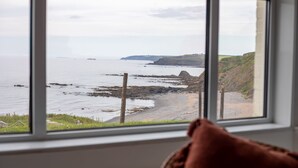 Vista da janela mostrando uma paisagem costeira com costa rochosa, uma colina gramada e um navio ao longe na água.