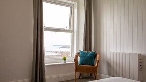 Quarto de hotel com cama, uma cadeira com almofada, janela com cortinas e uma pequena planta no parapeito.