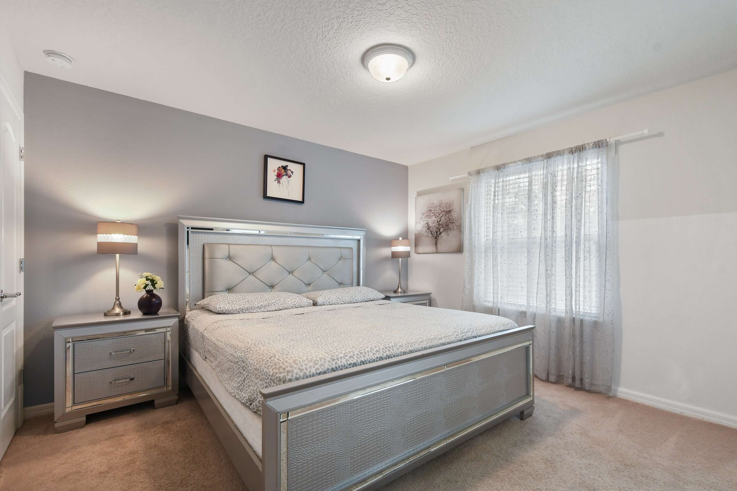Quarto com cama, duas mesinhas de cabeceira, um criado-mudo, luminária na parede e janela com cortinas finas.