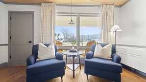 Quarto com duas poltronas azuis, uma mesa redonda pequena, um abajur e uma janela grande com cortinas.