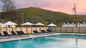 Uma piscina com espreguiçadeiras e guarda-sóis, uma cerca de madeira e uma encosta arborizada ao fundo.